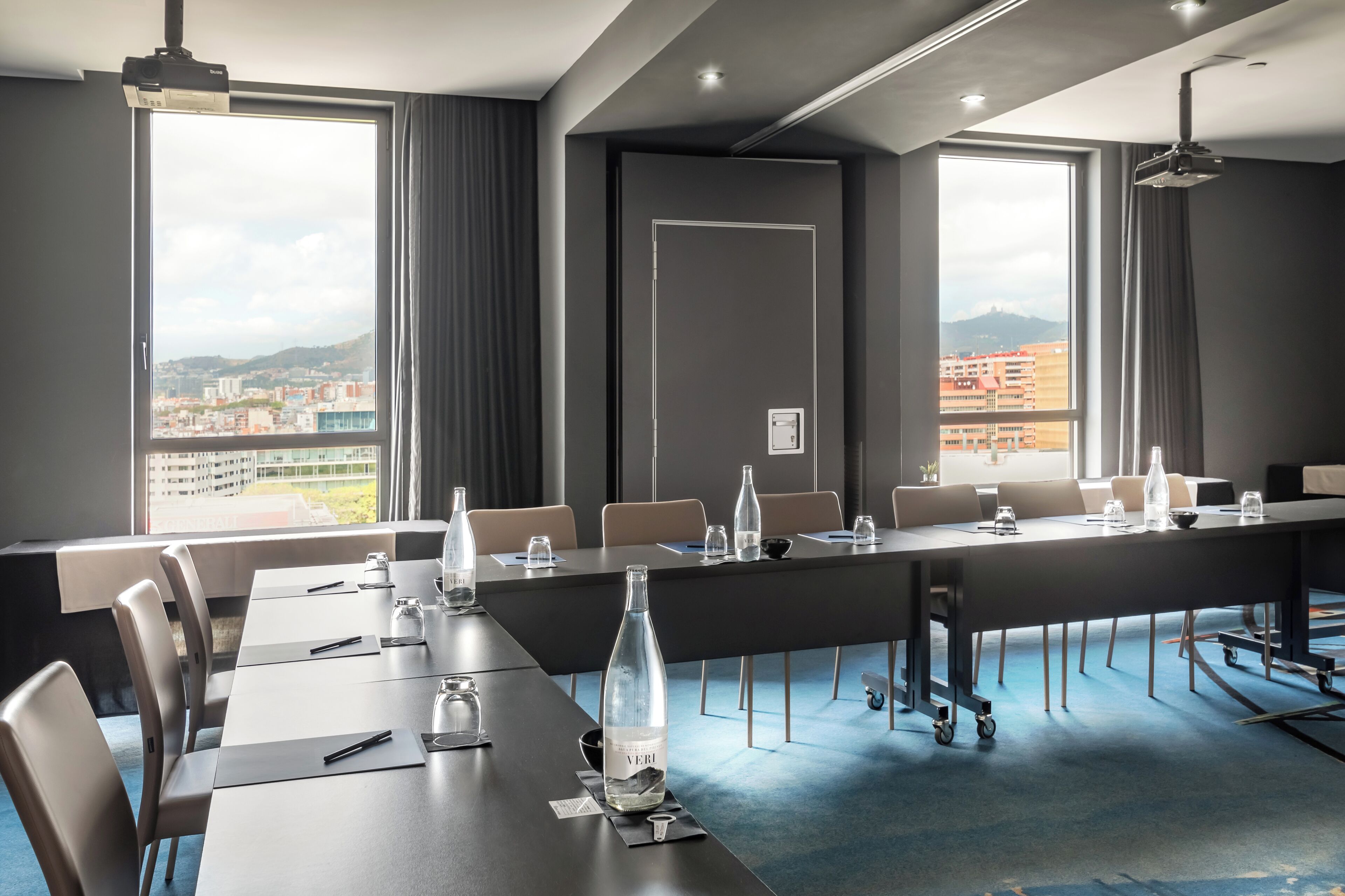Sala de conferências com uma mesa comprida, cadeiras, garrafas de água e vista para o horizonte urbano através de grandes janelas.