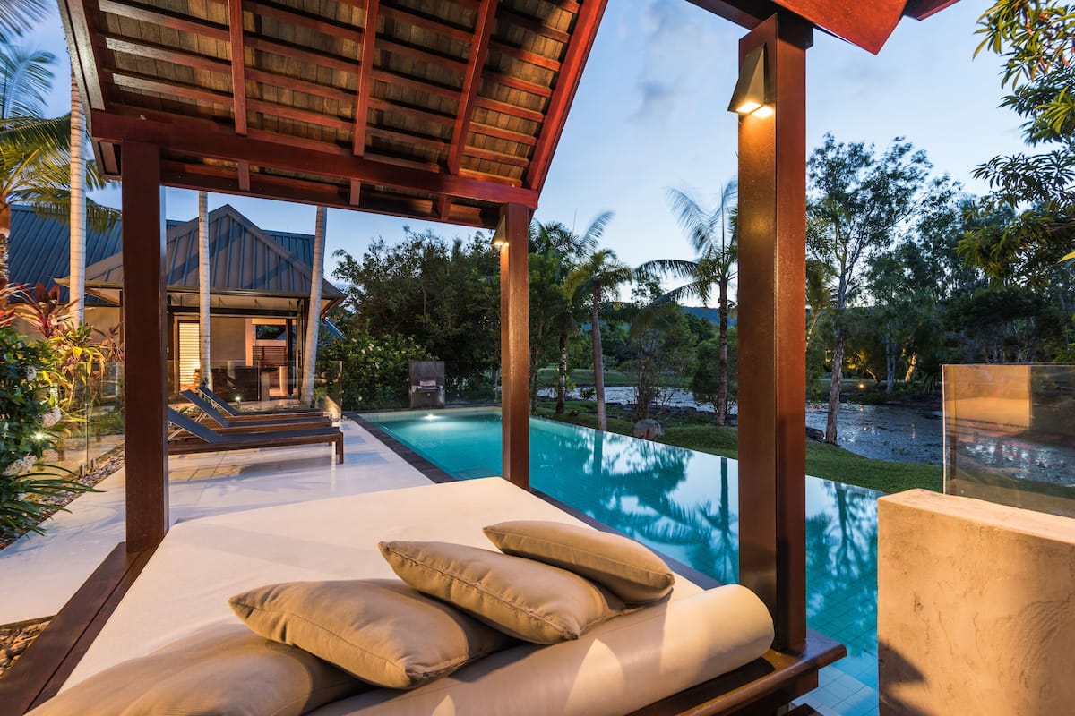 Área da piscina com um pérgola de madeira, cadeiras de descanso e vista para uma casa e piscina.