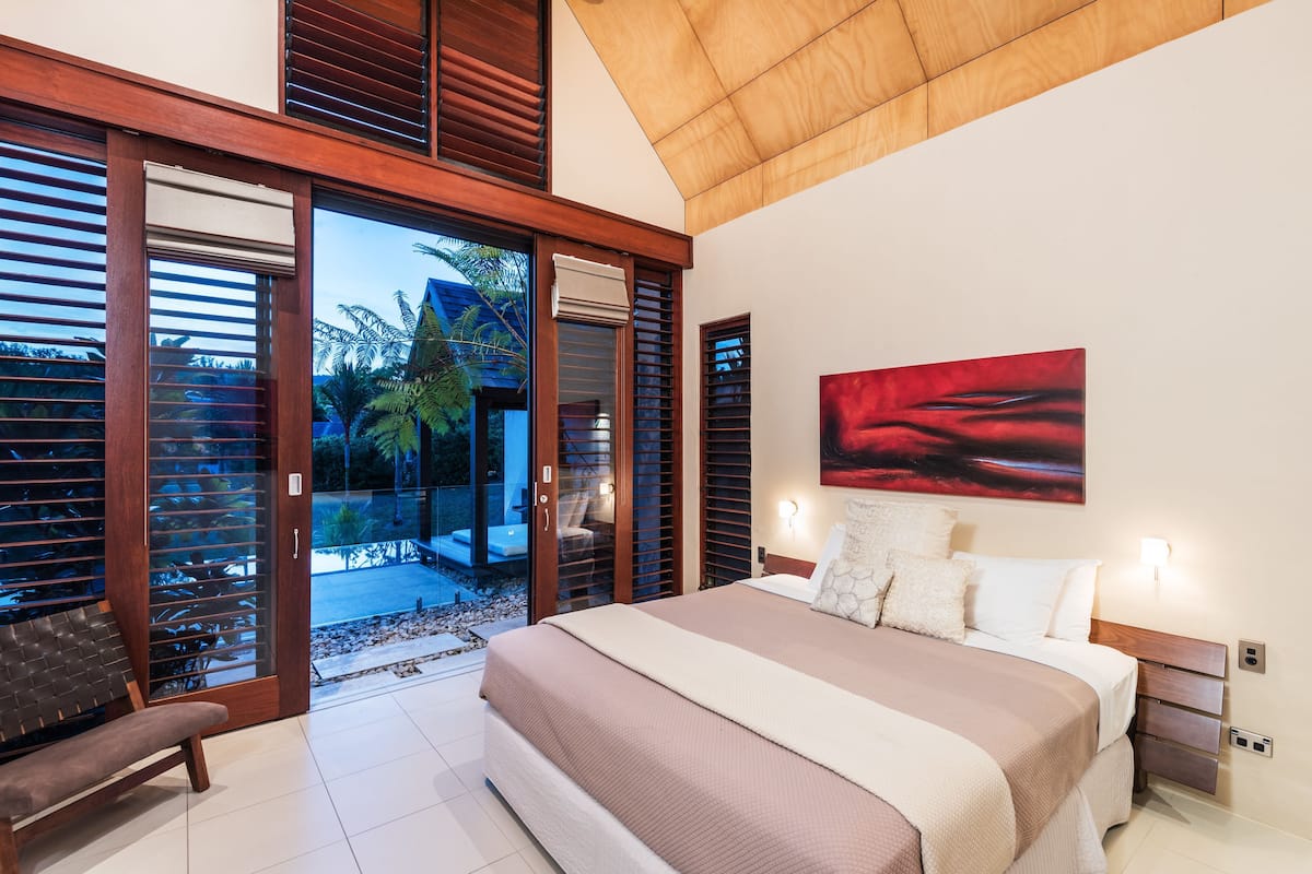 Quarto com cama, um banco, porta de vidro deslizante para piscina e um quadro abstrato vermelho.