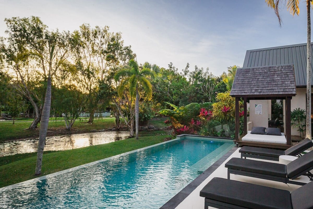 Piscina com espreguiçadeiras, cercada por vegetação exuberante e um edifício moderno.