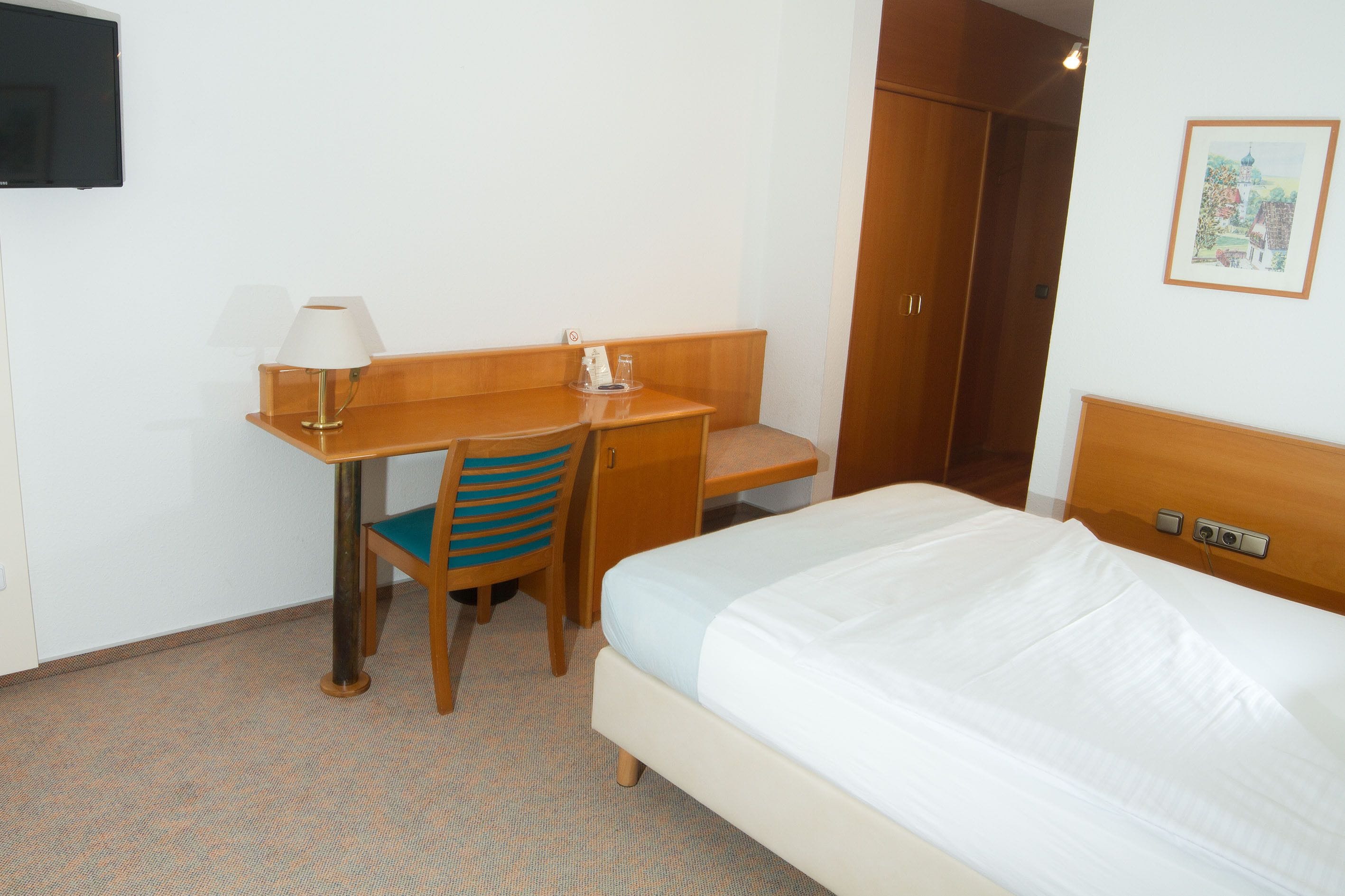 Quarto de hotel com cama, escrivaninha, cadeira, luminária e quadro na parede.