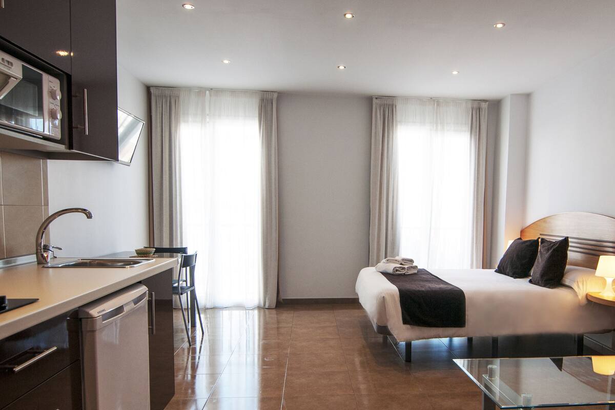 Quarto de hotel moderno com cama, mesa de cabeceira, luminária e área de kitchenette.