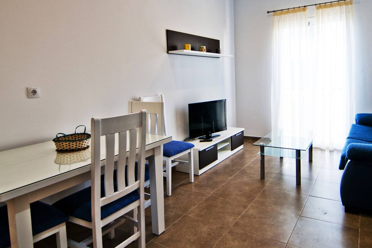 Uma sala de estar com televisão, um sofá azul, uma mesa de jantar com cadeiras e um cesto sobre a mesa.