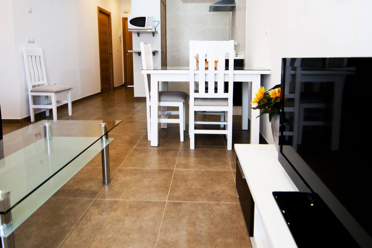 Uma sala de estar moderna com uma mesa de centro de vidro, mobiliário branco, uma TV de tela plana e um micro-ondas na parede.
