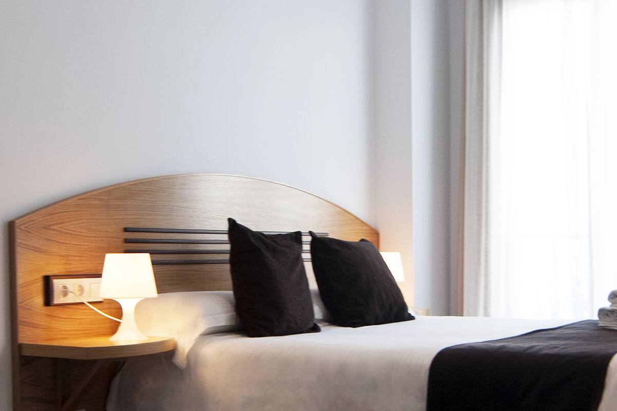 Quarto de hotel com cabeceira de madeira, duas almofadas pretas, colcha branca, abajur e janela com cortinas.