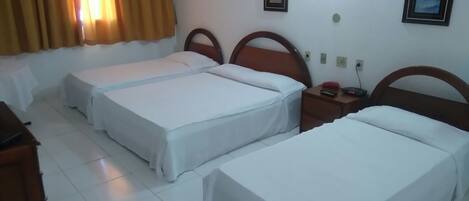Quarto de hotel com duas camas de solteiro, uma mesa de cabeceira de madeira com abajur e uma janela com cortinas.