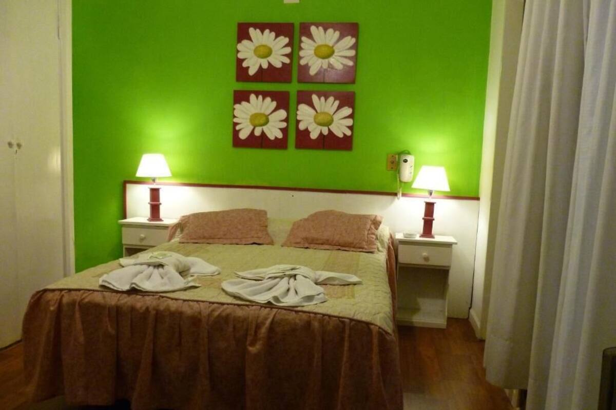 Cama bem arrumada com colcha marrom e duas almofadas, com mesas de cabeceira laterais com abajures.