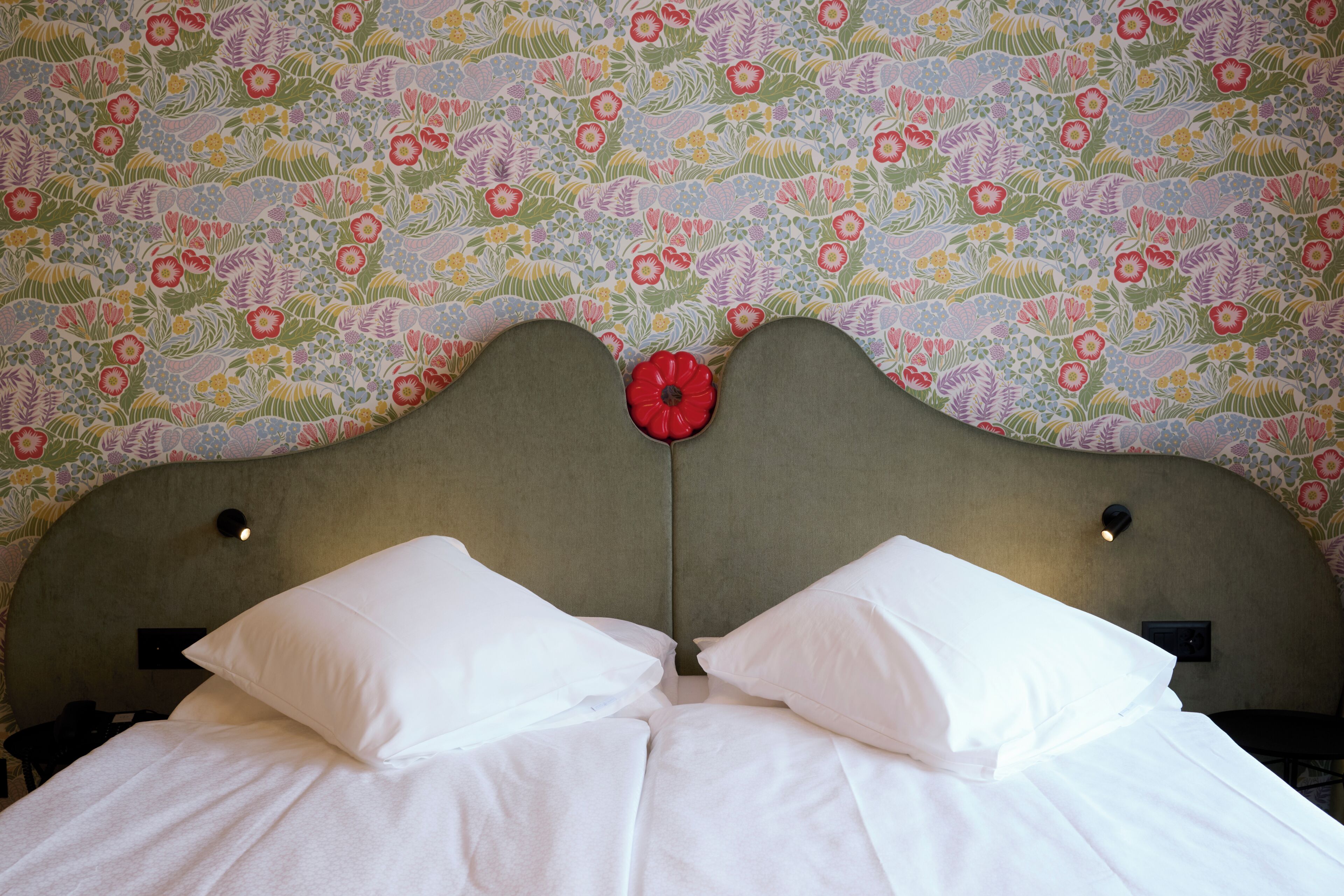 Uma cama com roupa de cama branca, cabeceira decorada com detalhes vermelhos e papel de parede estampado com flores.