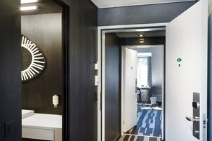 Quarto de hotel moderno com parede escura, piaça branca e porta que dá para um corredor com carpete estampado.