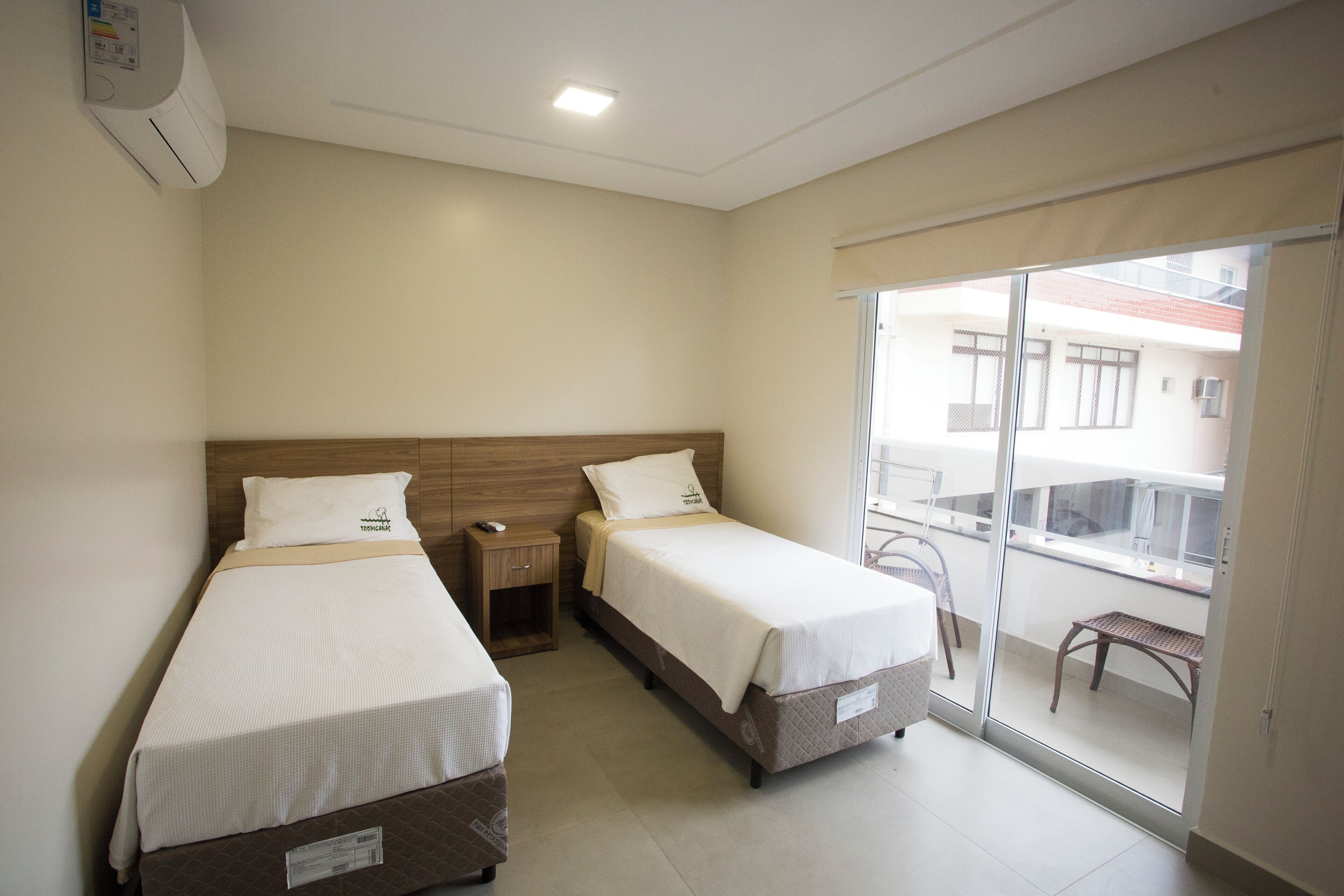 Quarto de hotel com duas camas, uma mesa de cabeceira com controle remoto, uma janela com vista para prédios e uma varanda com uma cadeira.