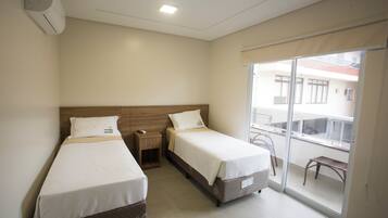 Quarto de hotel com duas camas, uma mesa de cabeceira com controle remoto, uma janela com vista para prédios e uma varanda com uma cadeira.