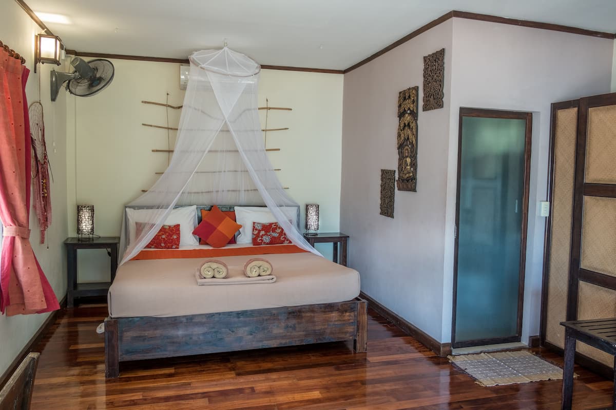 Quarto com cama, mesa de cabeceira, ventoinha, mosquiteiro e uma porta.