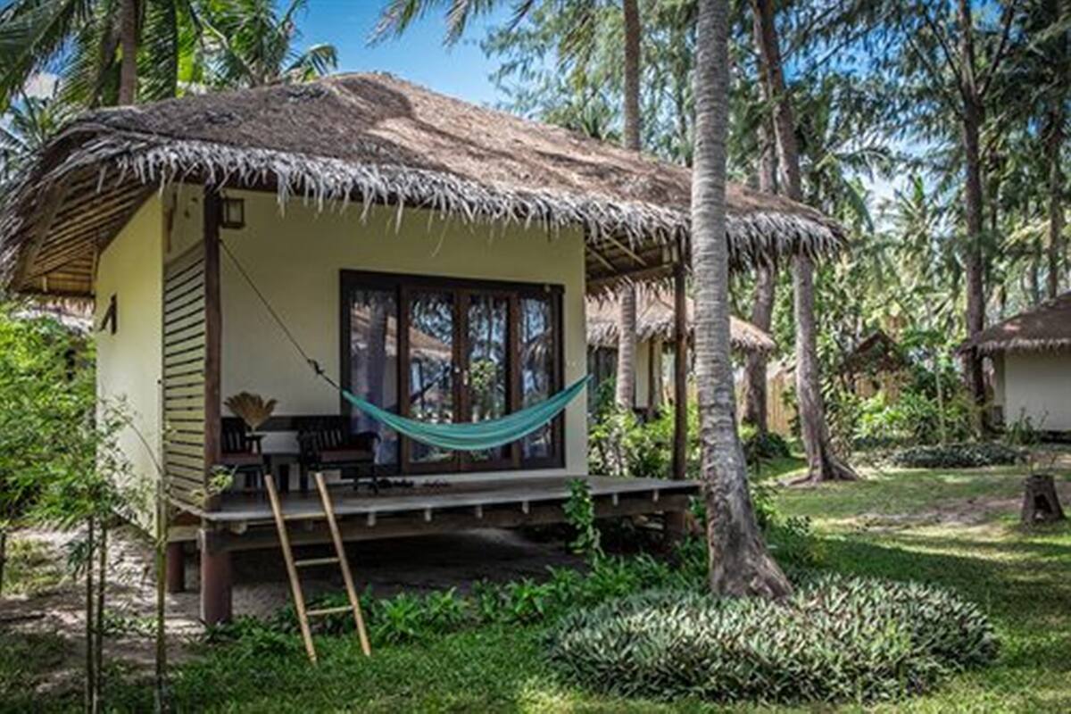 Uma cabana com telhado de palha e varanda, rodeada por vegetação exuberante e palmeiras.