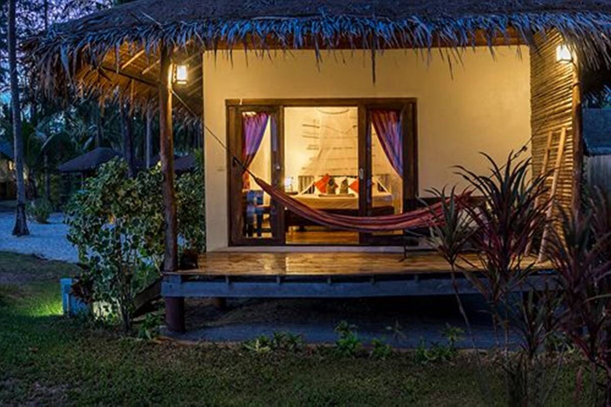 Estrutura com telhado de palha, com rede e um deck de madeira, cercada por vegetação tropical.