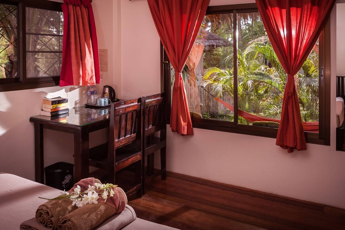 Quarto com escrivaninha de madeira, uma cadeira, uma cama e janela com cortinas vermelhas.
