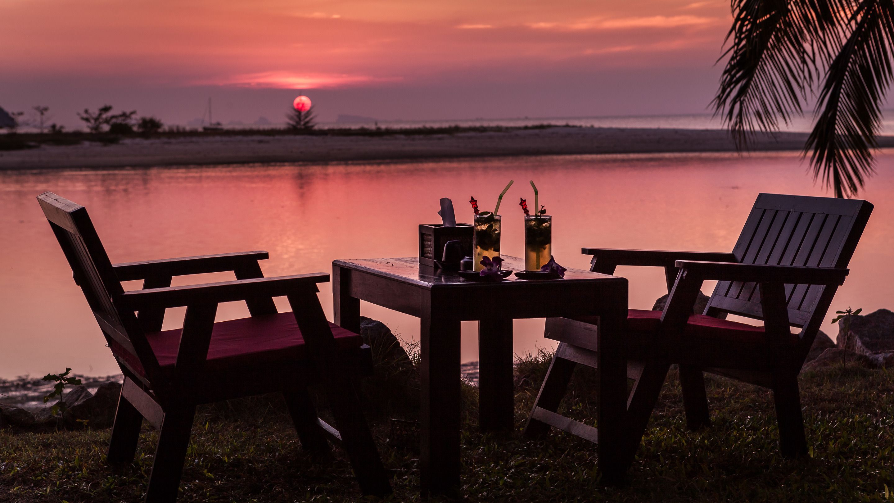 Vista do pôr do sol com uma mesa e cadeiras à beira da água.