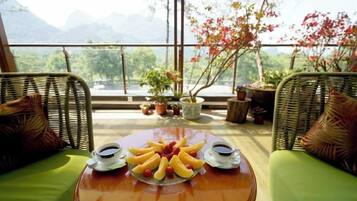 Terraço com móveis de vime, uma mesa com frutas e bebidas, e vista para montanhas.