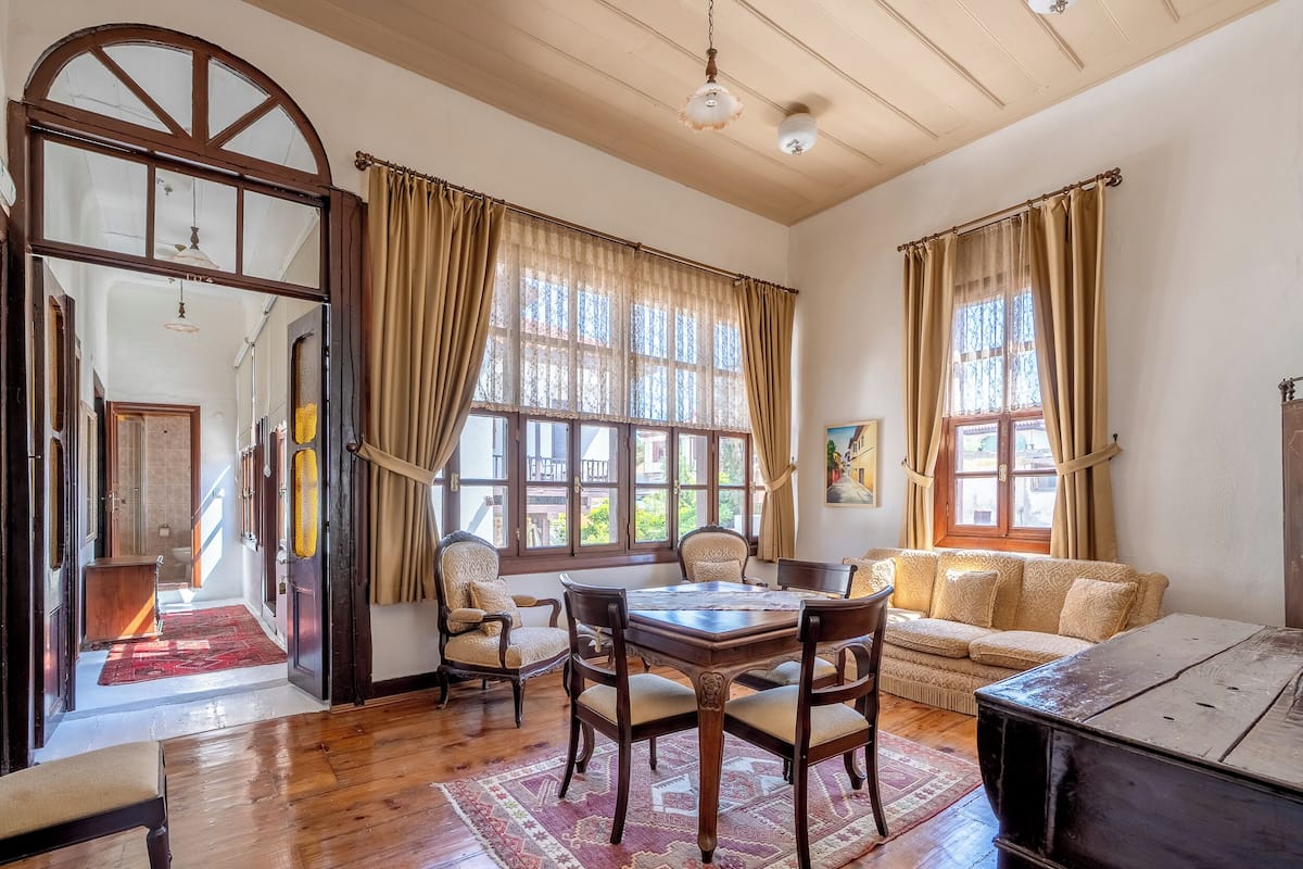 Um quarto com uma mesa de jantar de madeira, cadeiras, um sofá e uma janela grande com cortinas.