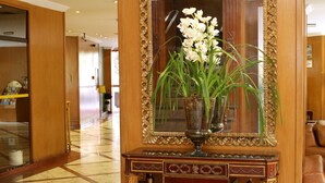 Um hall com piso de mármore, balcão de recepção de madeira, um espelho e um vaso com flores brancas.