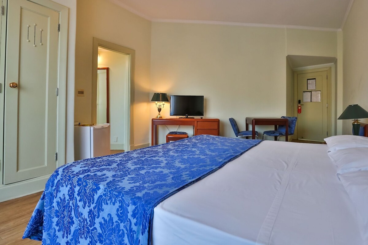Quarto de hotel com uma cama, escrivaninha com televisão, uma cadeira e guarda-roupa.