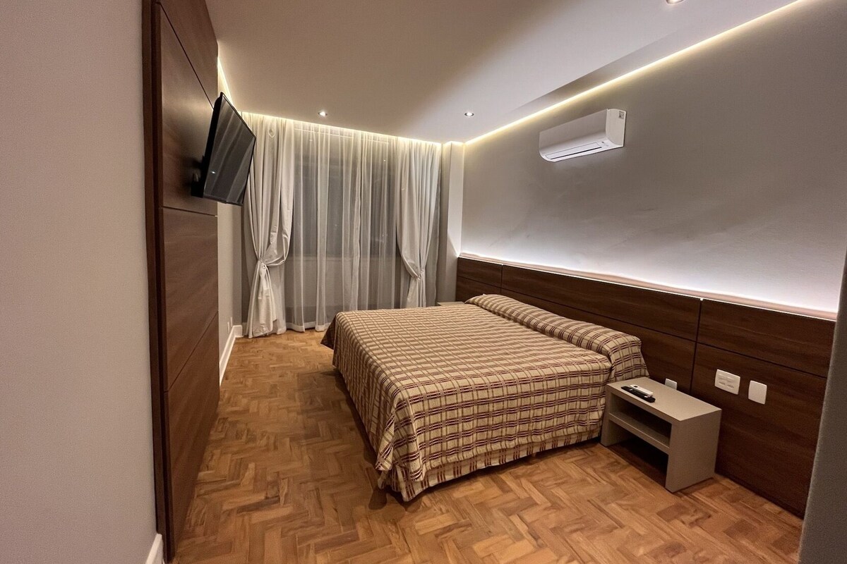 Quarto de hotel moderno com piso de madeira em padrão herringbone, cama com roupa de cama xadrez, mesa de cabeceira, televisão montada na parede e ar condicionado.