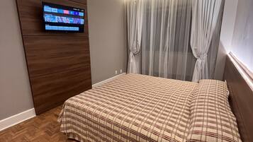 Quarto moderno com cabeceira de madeira, televisão de tela plana fixada nela, cama com colcha xadrez, mesa de cabeceira com controle remoto e cortinas translúcidas na janela.