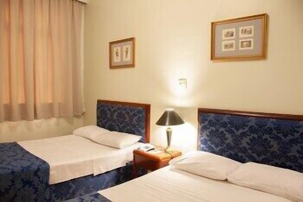 Quarto de hotel com duas camas, mesa de cabeceira com abajur e quadros nas paredes.