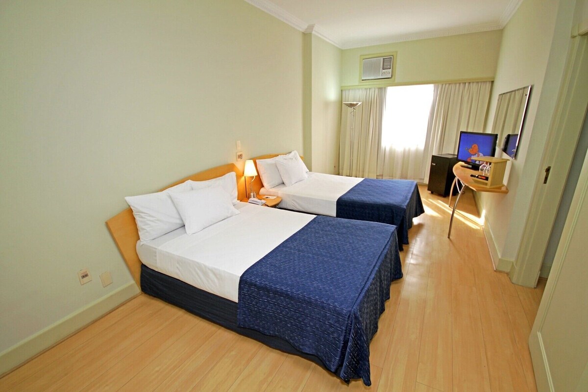 Quarto de hotel com duas camas, uma escrivaninha com computador, televisão e janela com cortinas.