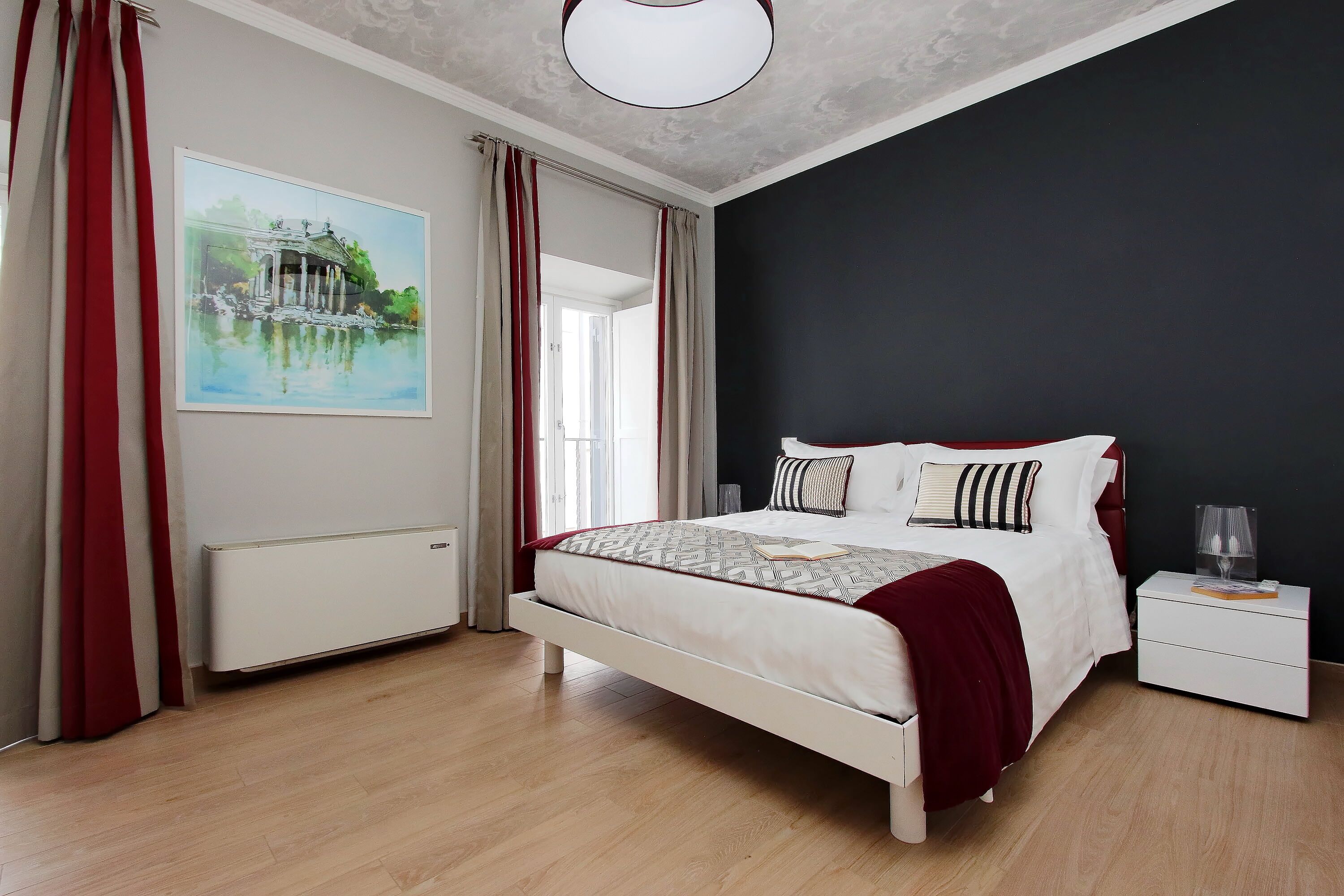 Um quarto com cama, mesas de cabeceira, um criado-mudo, um quadro e uma janela com cortinas.