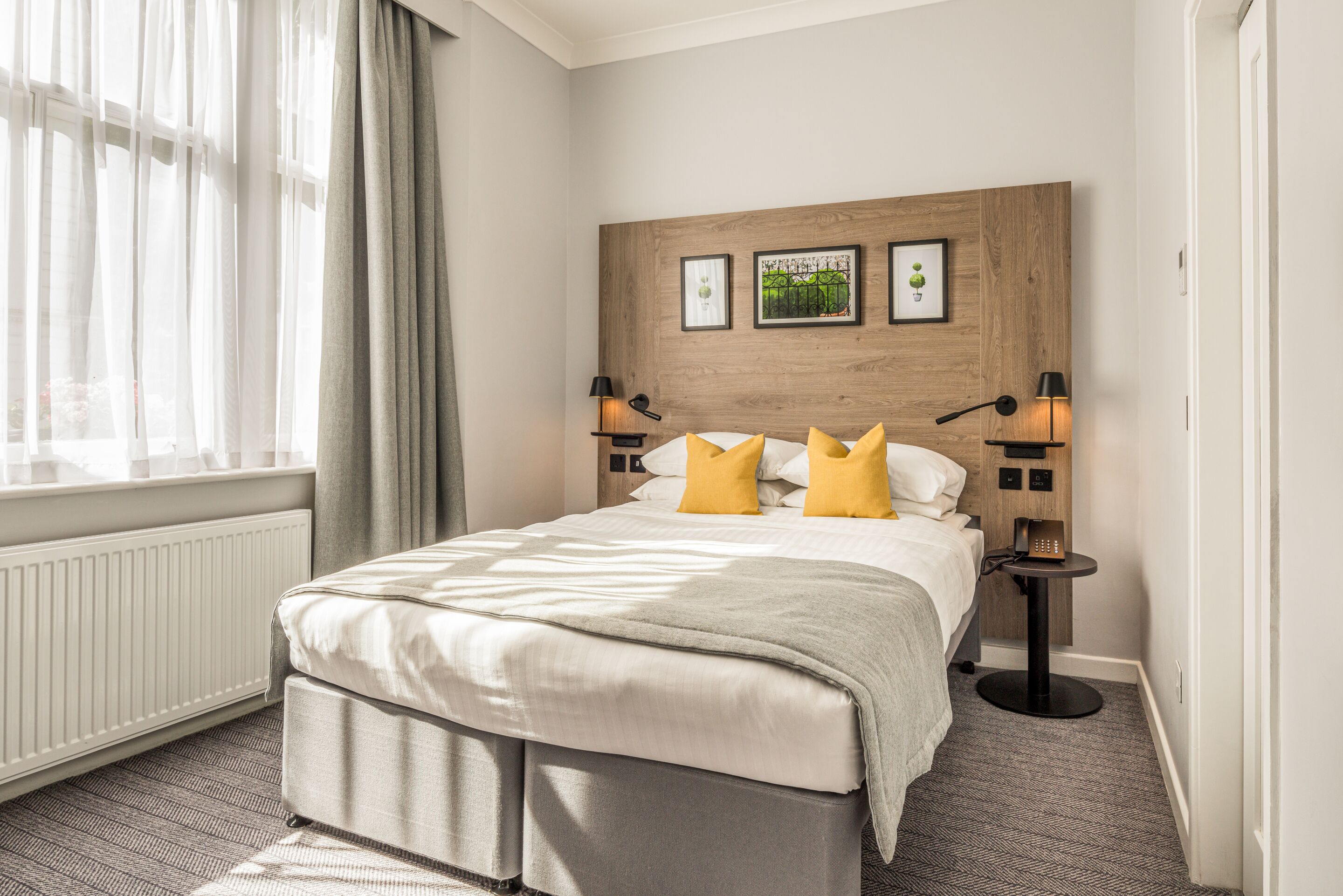 Quarto de hotel com cama, luminárias de cabeceira, cabeceira com quadros e uma janela com cortinas, além de um radiador.