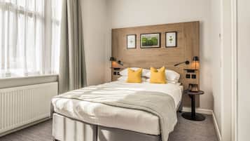 Quarto de hotel com cama, luminárias de cabeceira, cabeceira com quadros e uma janela com cortinas, além de um radiador.