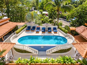Piscina com espreguiçadeiras e guarda-sóis, cercada por vegetação e edifícios.