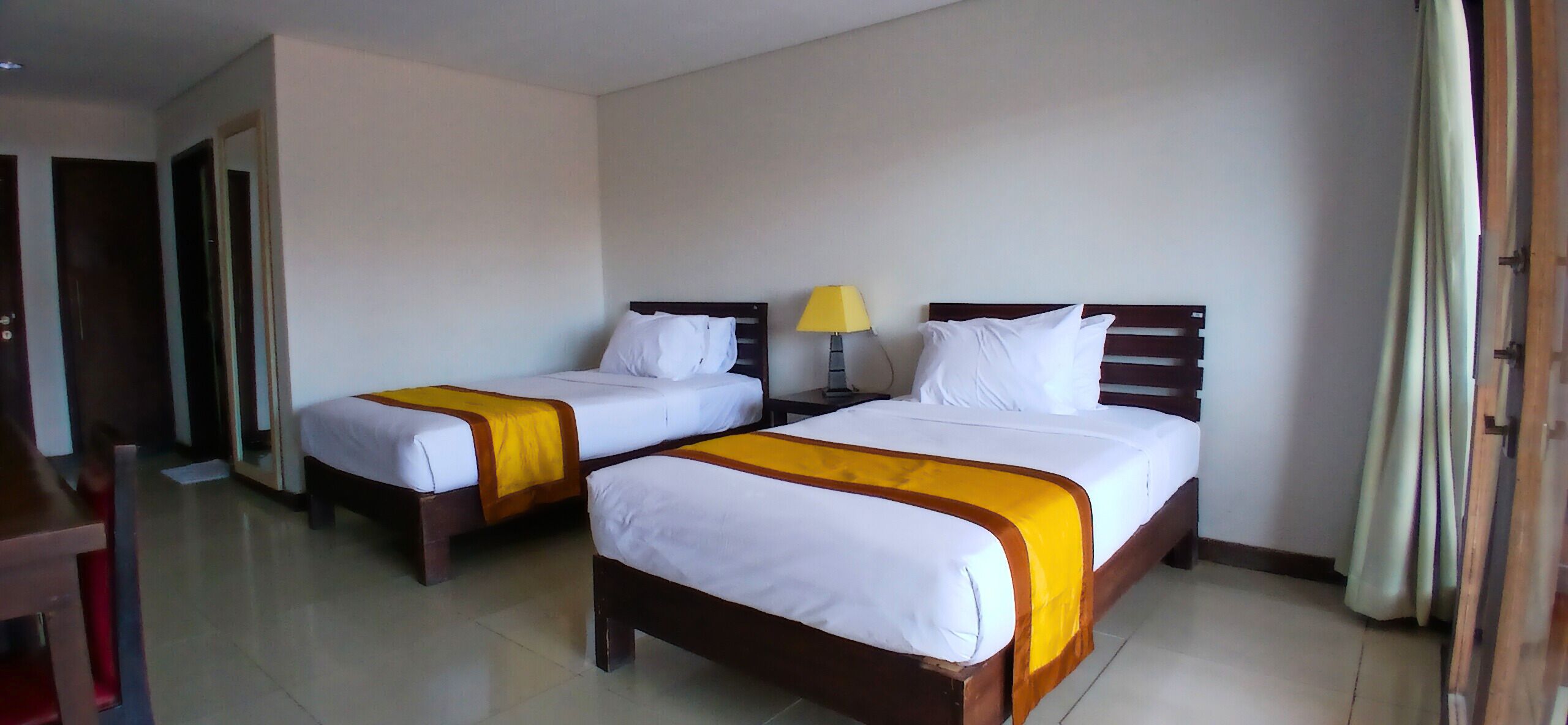 Quarto de hotel com duas camas, cada uma com cabeceira de madeira e roupa de cama branca. Há um abajur amarelo em uma das camas.