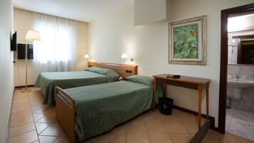 Quarto de hotel com duas camas, uma escrivaninha, luminária, pintura e banheiro.