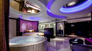 Um hall de hotel moderno com uma grande banheira de mármore, um espelho amplo e uma área de estar com um sofá roxo e uma mesa de centro preta.