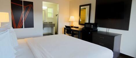 Quarto de hotel com cama, escrivaninha com luminária, televisão e espelho.