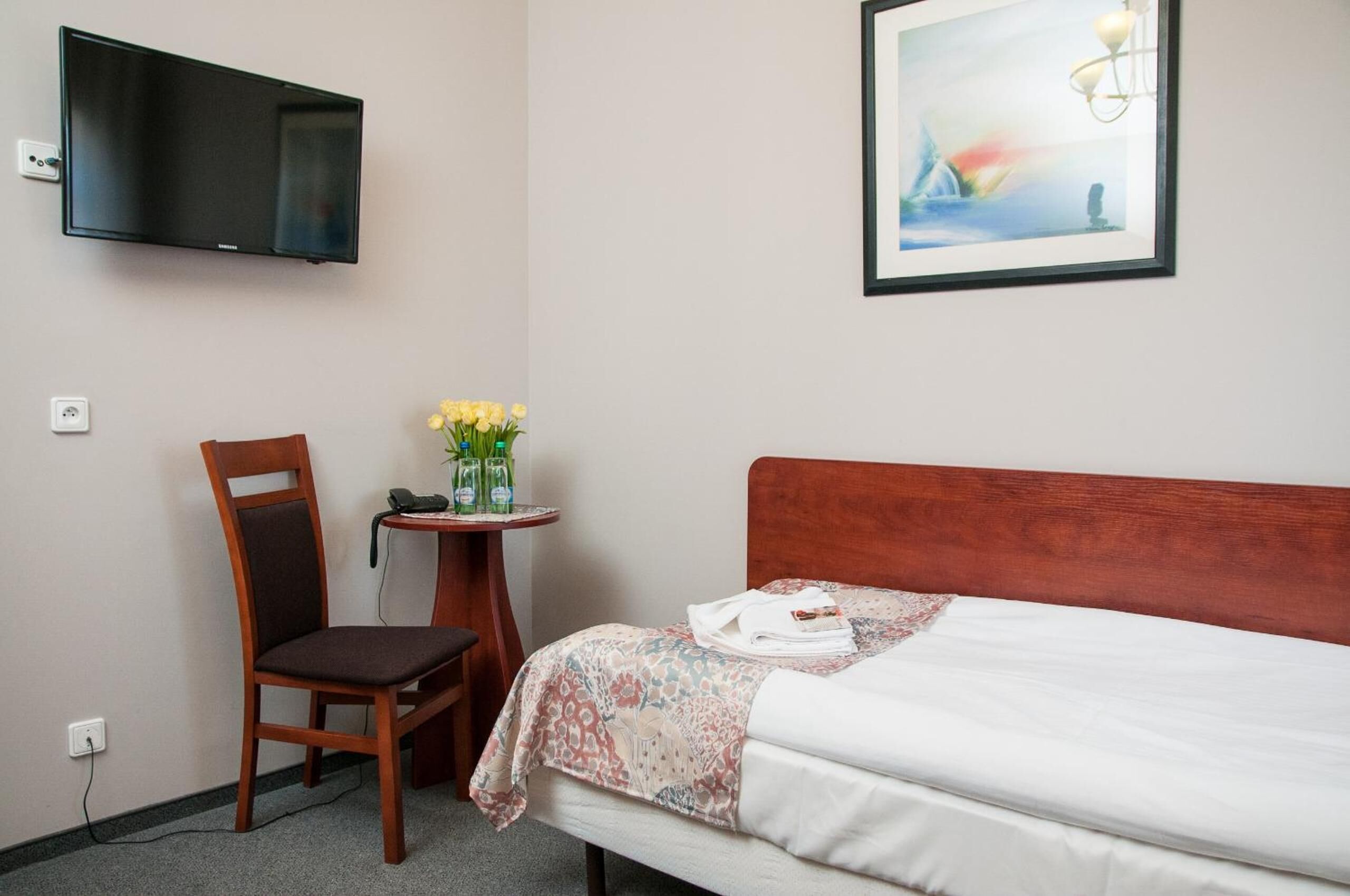 Quarto de hotel com uma cama, uma cadeira, uma pequena mesa com um vaso de flores, um quadro na parede e uma televisão montada acima da cama.