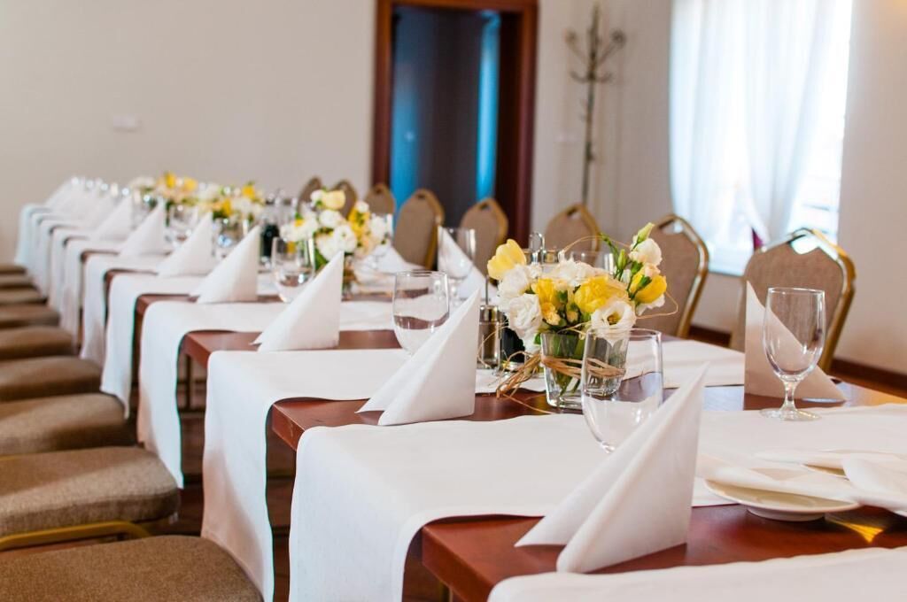 Uma sala de jantar de hotel com mesas compridas preparadas para o serviço, cada uma com toalha branca, copos e arranjos florais.