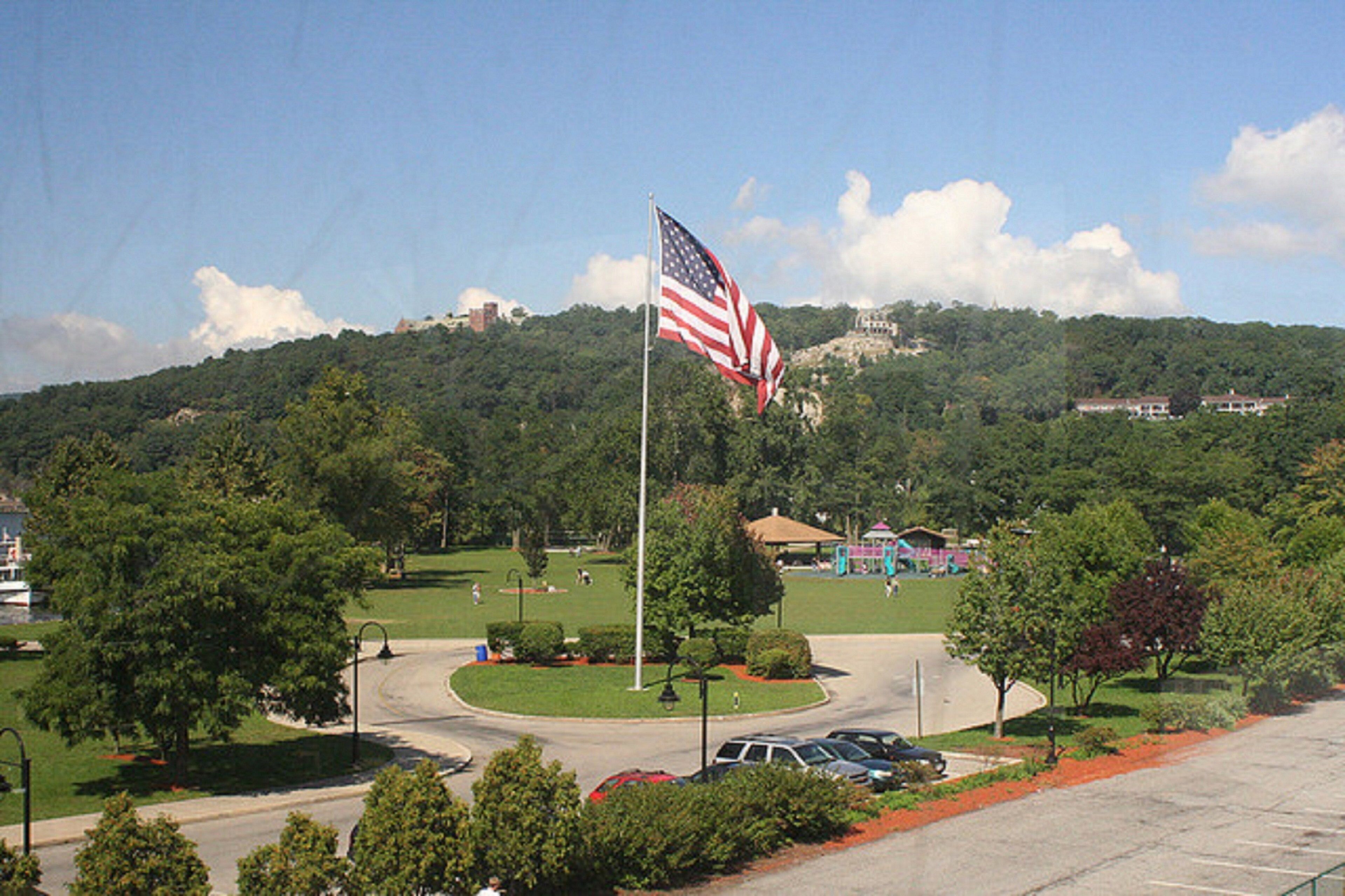 Um mastro com a bandeira americana, um estacionamento e uma área verde com árvores e um parquinho.