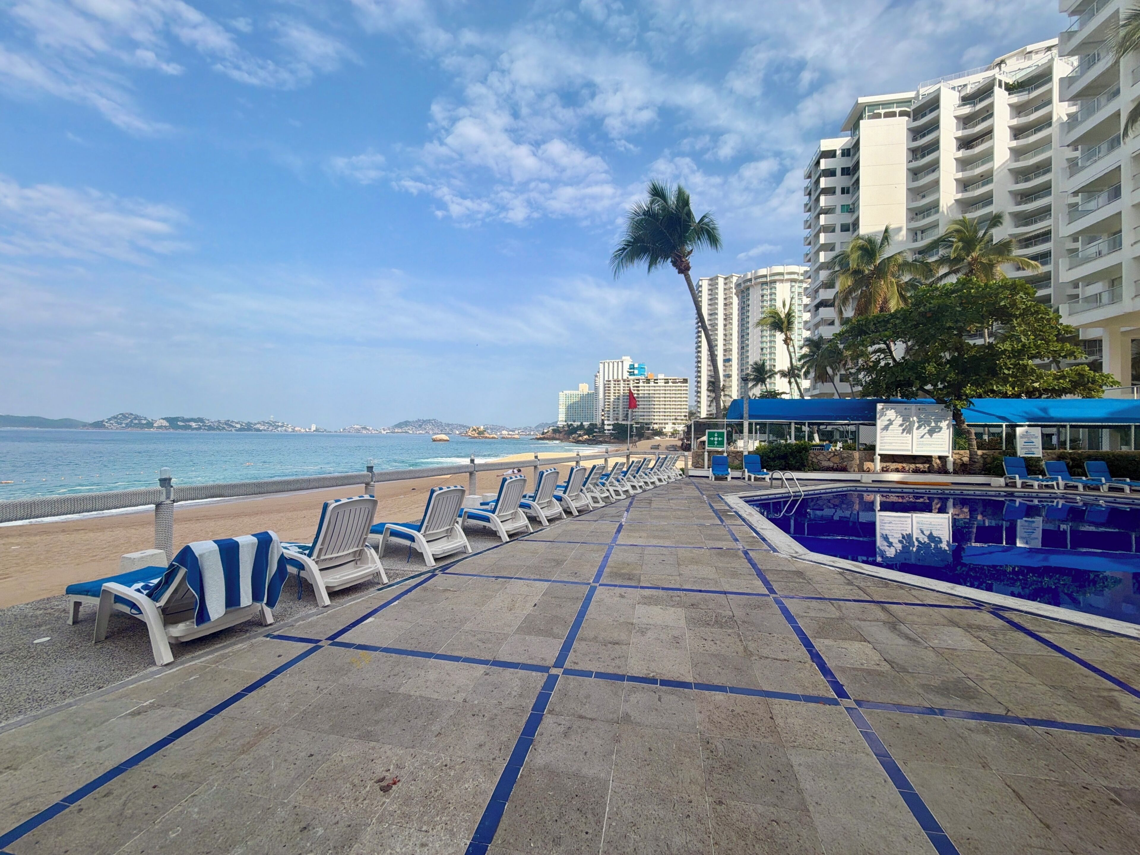 Área de piscina com espreguiçadeiras e piscina, com vista para uma praia e céu claro.
