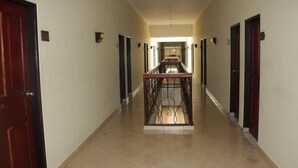 Corredor longo e estreito de um hotel, com corrimão central, portas em ambos os lados e piso de azulejos.