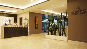 Uma área de recepção com balcão, dois funcionários, um arranjo decorativo de orquídeas e uma parede com obra de arte cultural.