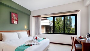 Quarto de hotel com cama, escrivaninha e uma grande janela com vista para árvores.