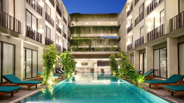 Piscina com espreguiçadeiras, cercada por um edifício com várias andares, varandas e vegetação.