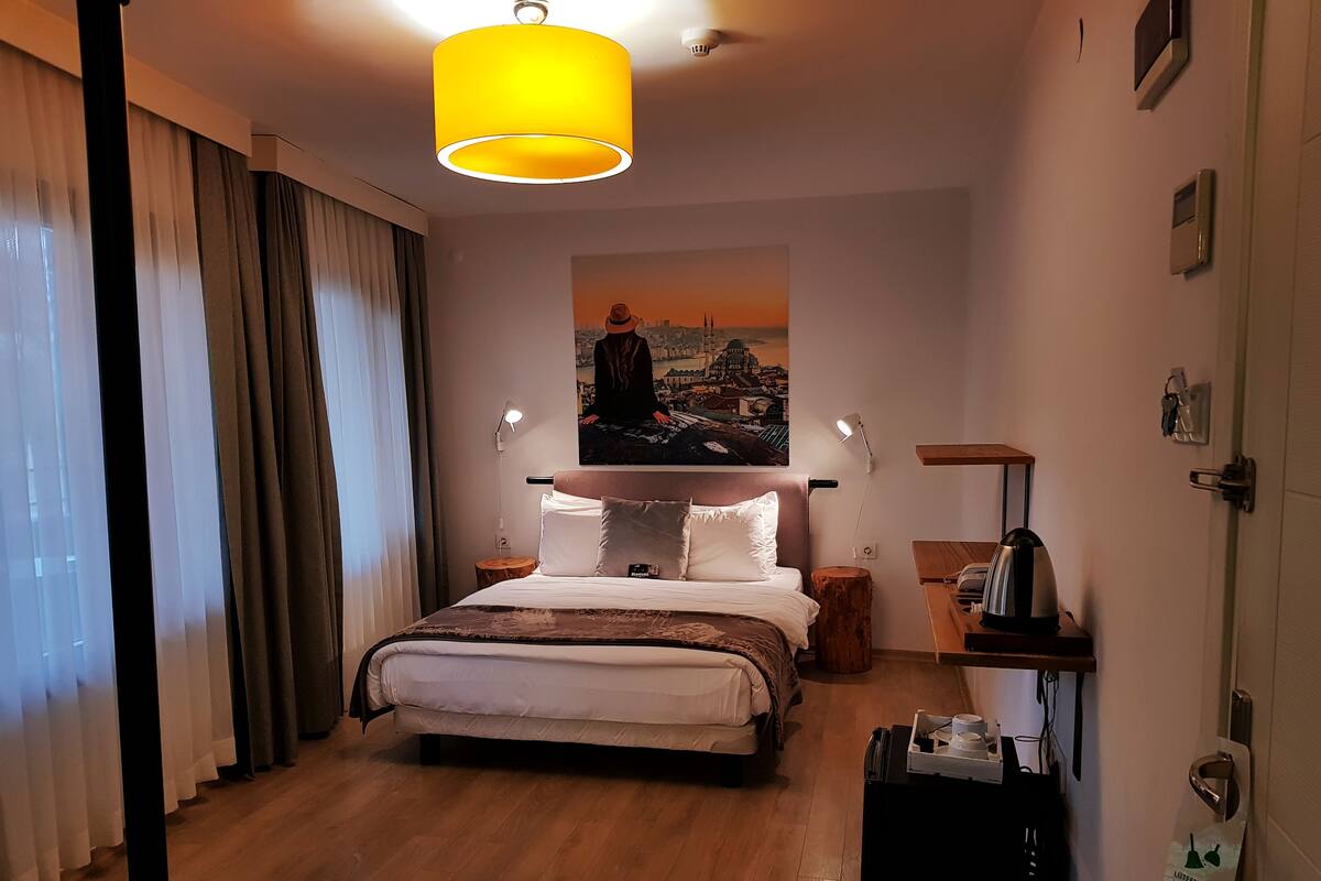 Quarto de hotel com cama, mesa de cabeceira, luminária, quadro e cafeteira.