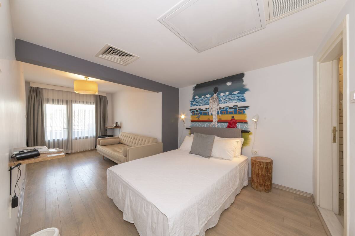 Quarto de hotel moderno com uma cama grande, um sofá, um quadro na parede e um banquinho de madeira.