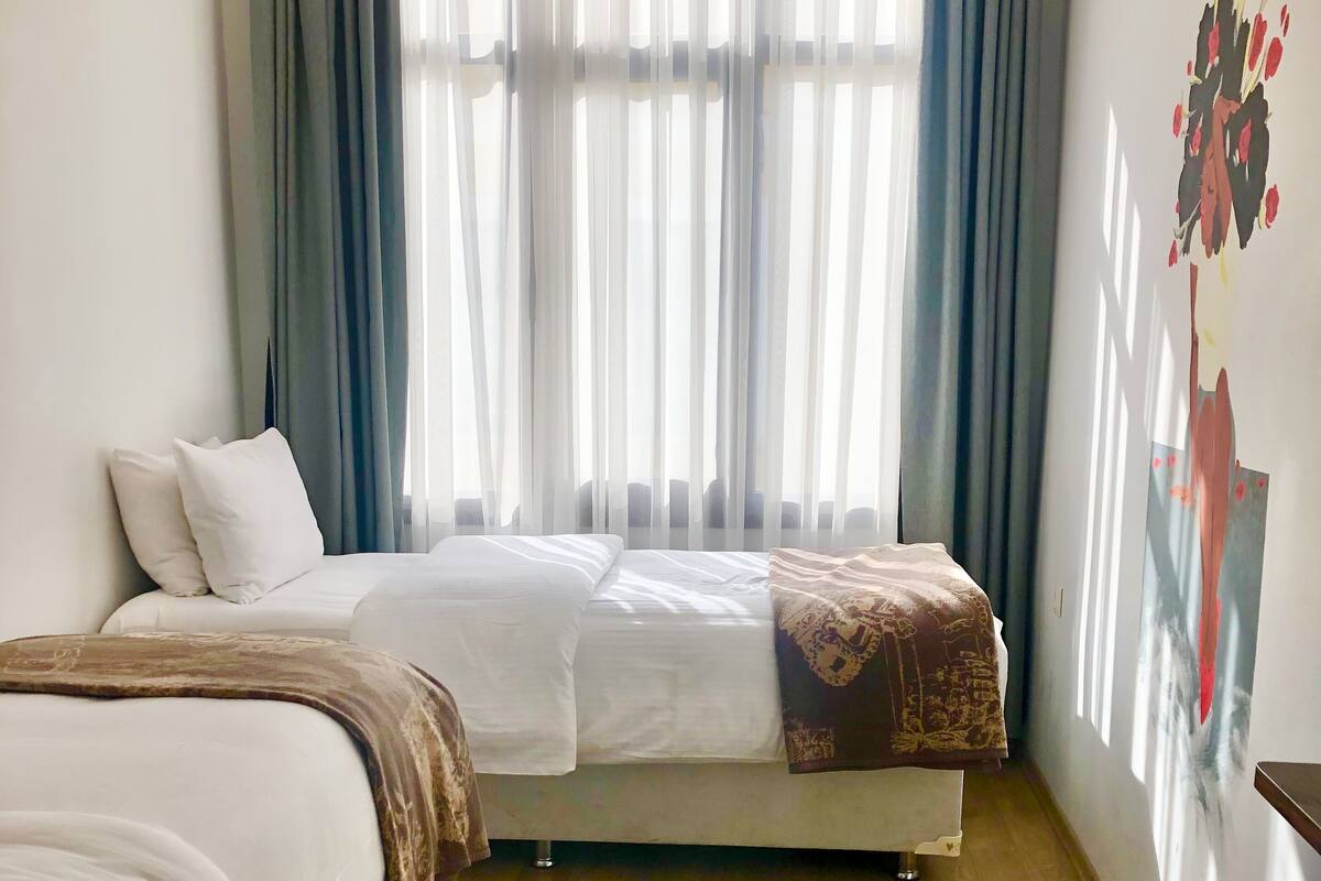 Quarto com duas camas, piso de madeira e uma janela grande com cortinas.