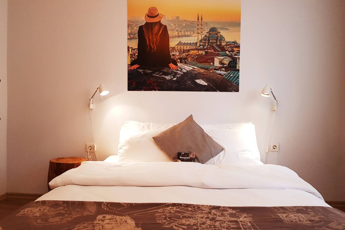 Cama com roupa de cama branca, um travesseiro e um cobertor. Tem uma mesa de cabeceira com uma luminária e um banquinho de madeira pequeno. Uma moldura com uma imagem de uma paisagem urbana está na parede acima da cama.