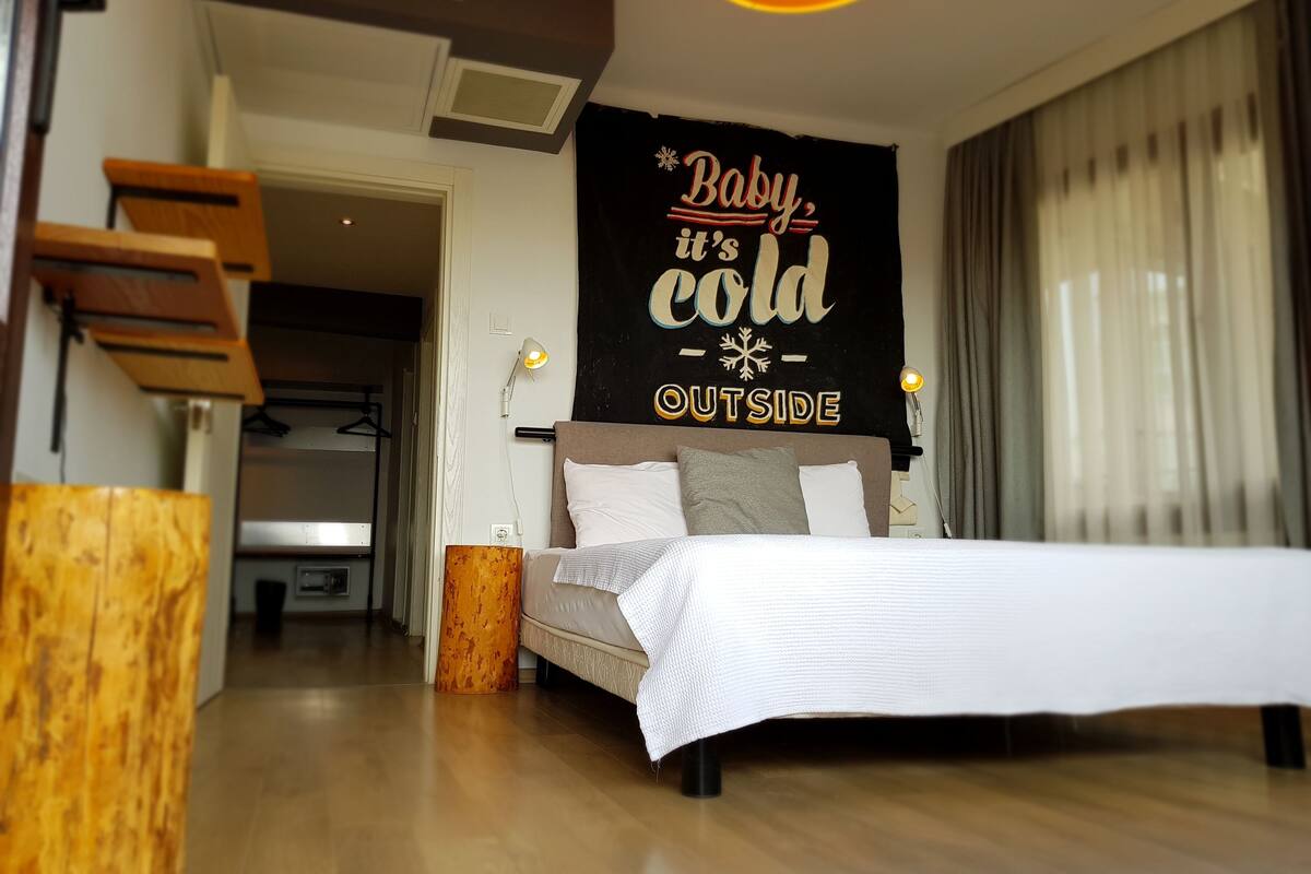 Quarto de hotel moderno com cama, luminárias de cabeceira, um letreiro fixado na parede e piso de madeira.