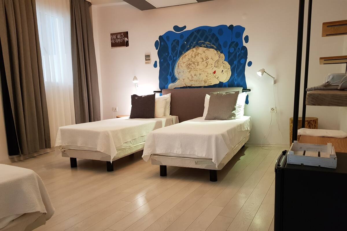 Quarto de hotel com duas camas de solteiro, uma grande obra de arte abstrata na parede, piso de madeira e uma pequena escrivaninha com luminária.